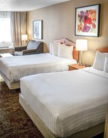 Quarto de hotel com duas camas, uma cadeira, escrivaninha, luminária e um quadro na parede.