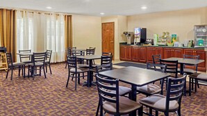 Uma sala de jantar de hotel com várias mesas e cadeiras, uma área de cozinha com eletrodomésticos e uma porta que dá para outro espaço.