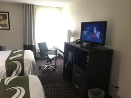 Quarto de hotel com duas camas, uma escrivaninha com computador, uma cadeira e televisão exibindo um anúncio.