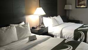 Quarto de hotel com duas camas, cada uma com roupa de cama branca e detalhes verdes, uma mesa de cabeceira com abajur, um relógio de parede e uma moldura com imagem.