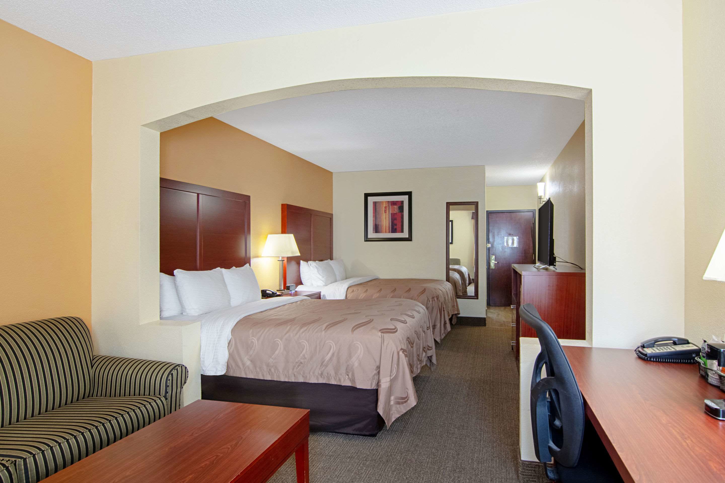 Quarto de hotel com cama, escrivaninha, cadeira, sofá, luminária e televisão.