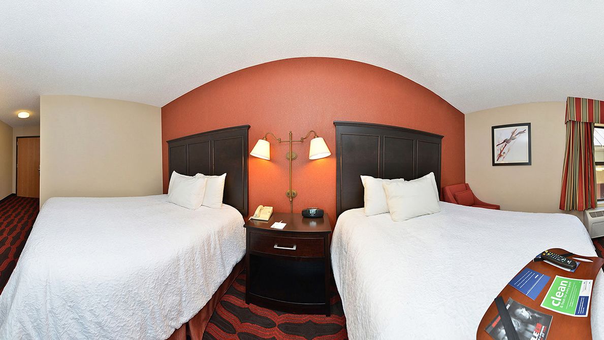 Quarto de hotel com duas camas, uma mesinha de cabeceira com telefone e abajur, e uma escrivaninha com controle remoto e folhetos.