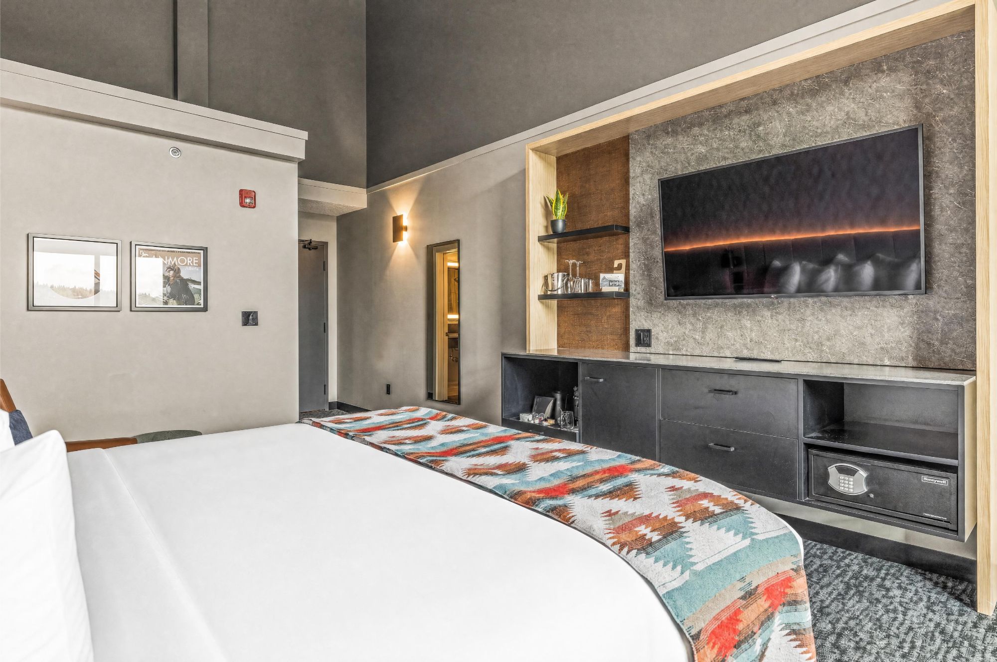 Quarto de hotel com cama, televisão presa a uma parede texturizada, minibar e porta que dá para outro cômodo.