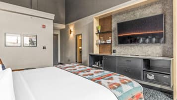 Quarto de hotel com cama, televisão presa a uma parede texturizada, minibar e porta que dá para outro cômodo.
