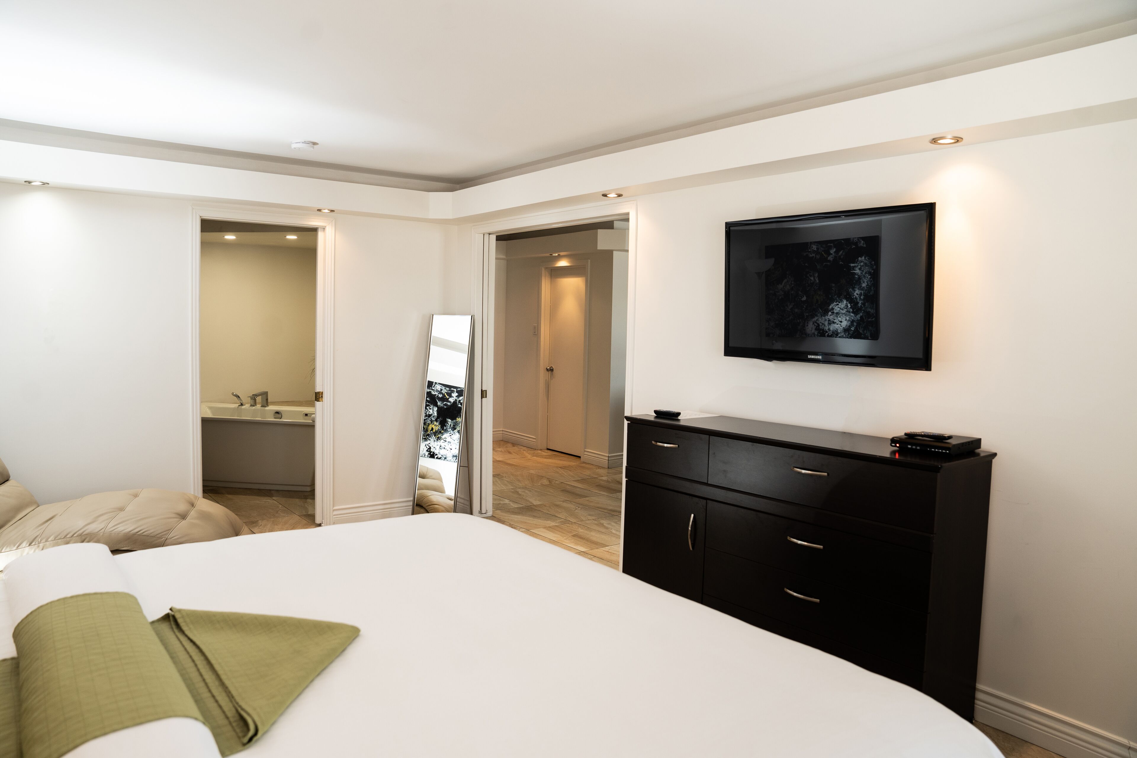 Quarto moderno com uma cama grande, cômoda preta, TV de tela plana e um banheiro visível por uma porta aberta.