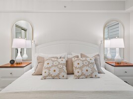 Quarto bem arrumado com cama branca, almofadas estampadas, duas mesas de cabeceira com luminárias e mesas de apoio com espelhos.