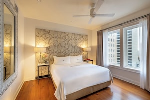 Quarto de hotel com uma cama grande, duas luminárias de cabeceira, um ventilador de teto e vista para edifícios através de grandes janelas.