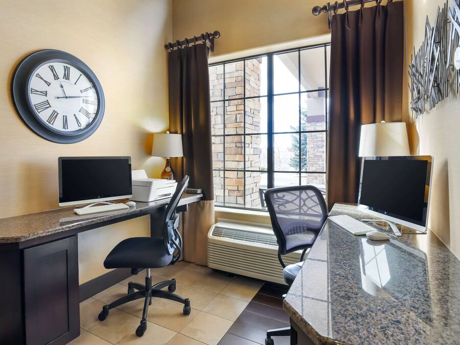 Escritório de hotel com duas mesas, computadores, cadeiras, um relógio e uma janela com cortinas.