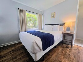 Uma cama bem arrumada com roupa de cama branca e azul marinho, chão de madeira, mesa de cabeceira com abajur e uma janela com cortinas.