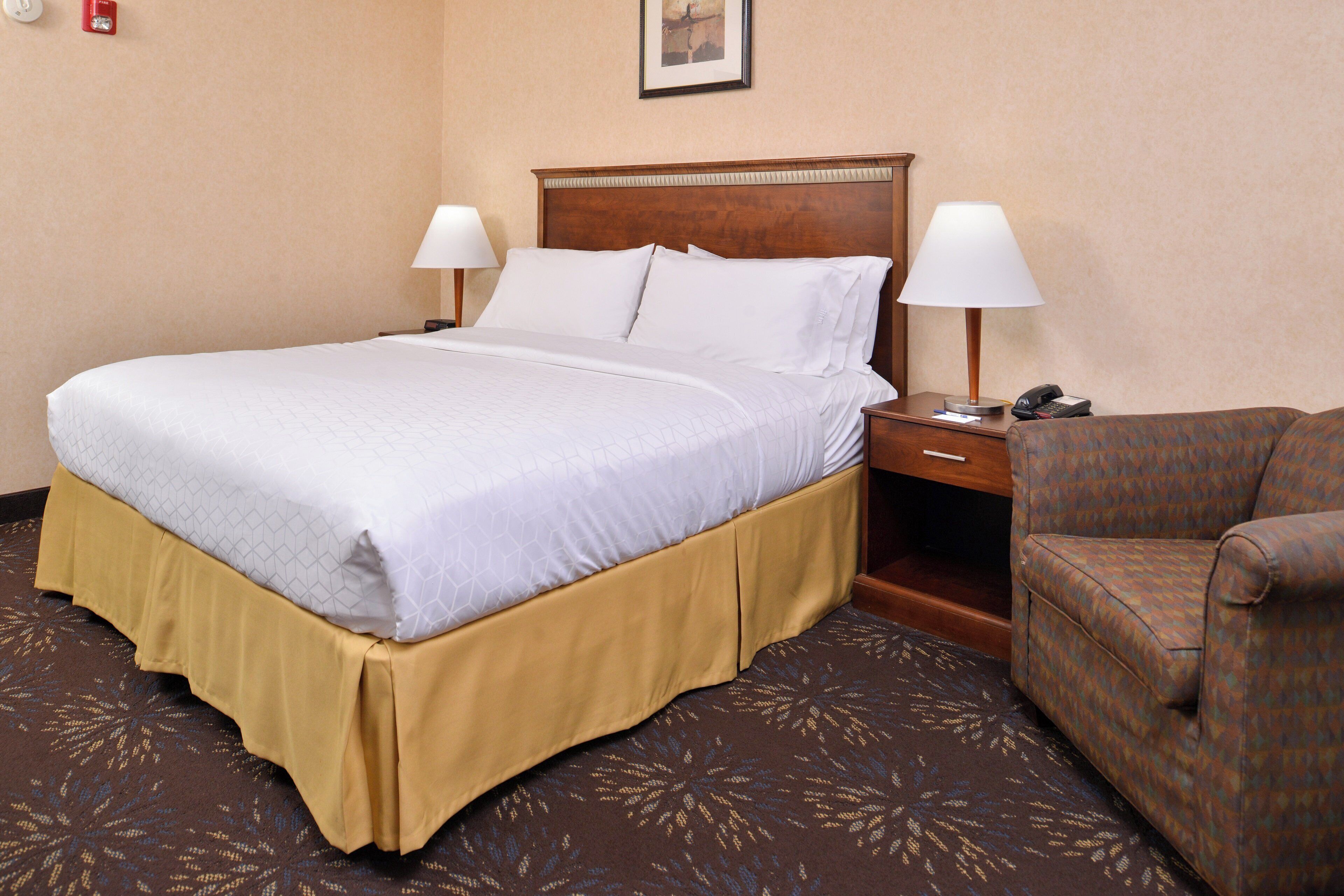 Quarto de hotel com cama, duas luminárias de cabeceira, telefone, cadeira e um quadro na parede.