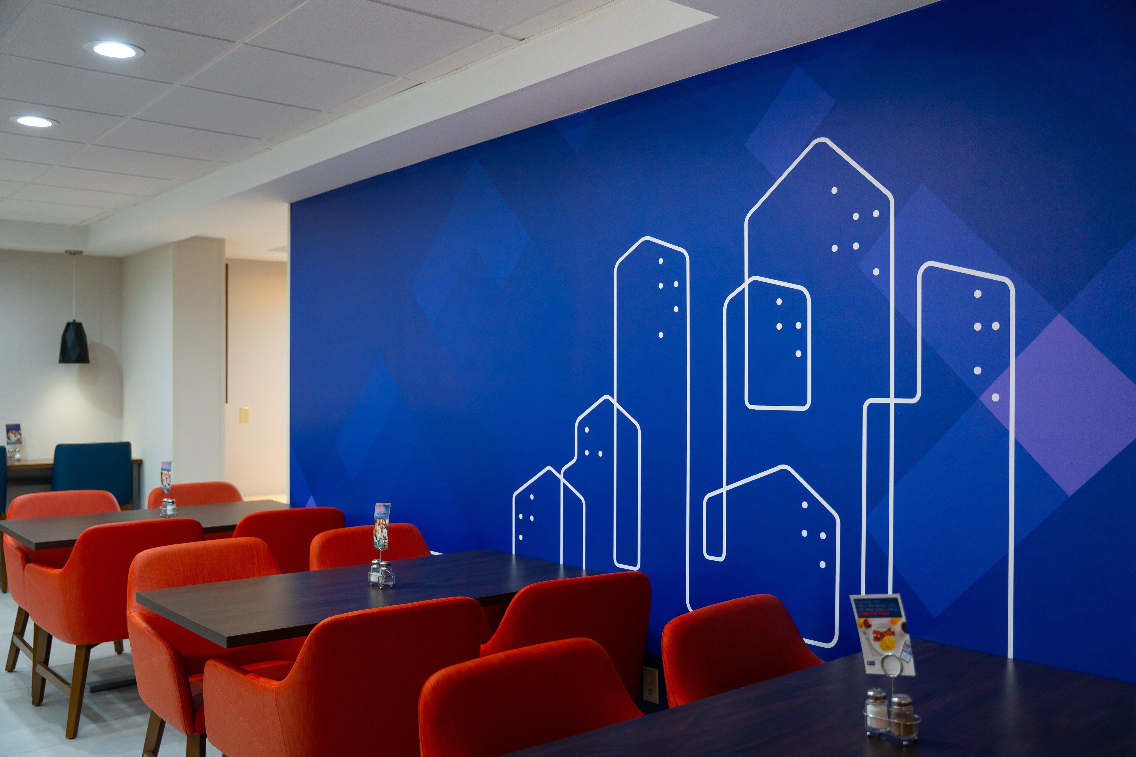 Sala de conferências com cadeiras vermelhas e uma mesa escura, apresentando uma parede azul com contornos de edifícios brancos.