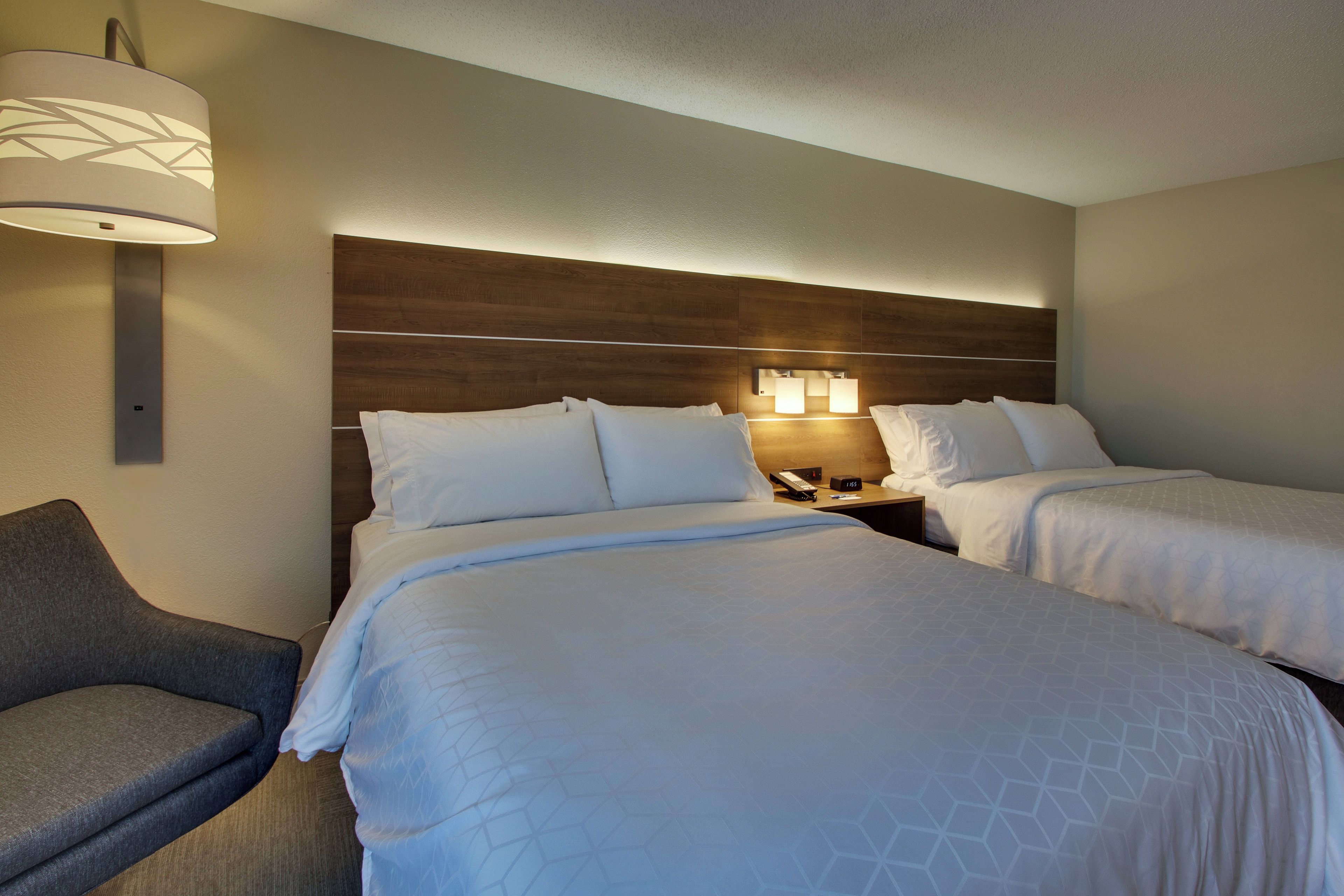 Quarto de hotel com duas camas, uma mesa de cabeceira com abajur e uma cadeira.