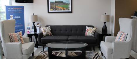 Uma sala de estar moderna com um sofá escuro, duas poltronas, uma mesa de centro de vidro e um quadro emoldurado na parede.