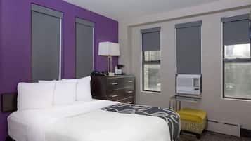 Quarto de hotel com cama, escrivaninha, luminária, janela e ar condicionado.