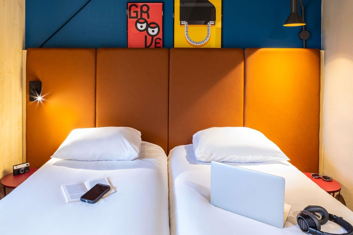 Quarto de hotel com duas camas, um laptop em uma cama, um par de fones de ouvido na outra e uma mesinha com abajur e óculos de sol.
