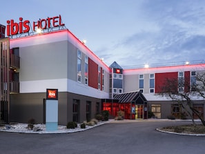 Um hotel moderno com um destaque para a placa 'ibis Hotel', uma entrada bem iluminada e uma área de estacionamento.
