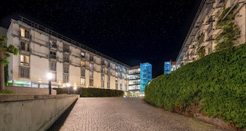 Vista noturna de um edifício com vários andares, um caminho de pedras e uma sebe bem cuidada.