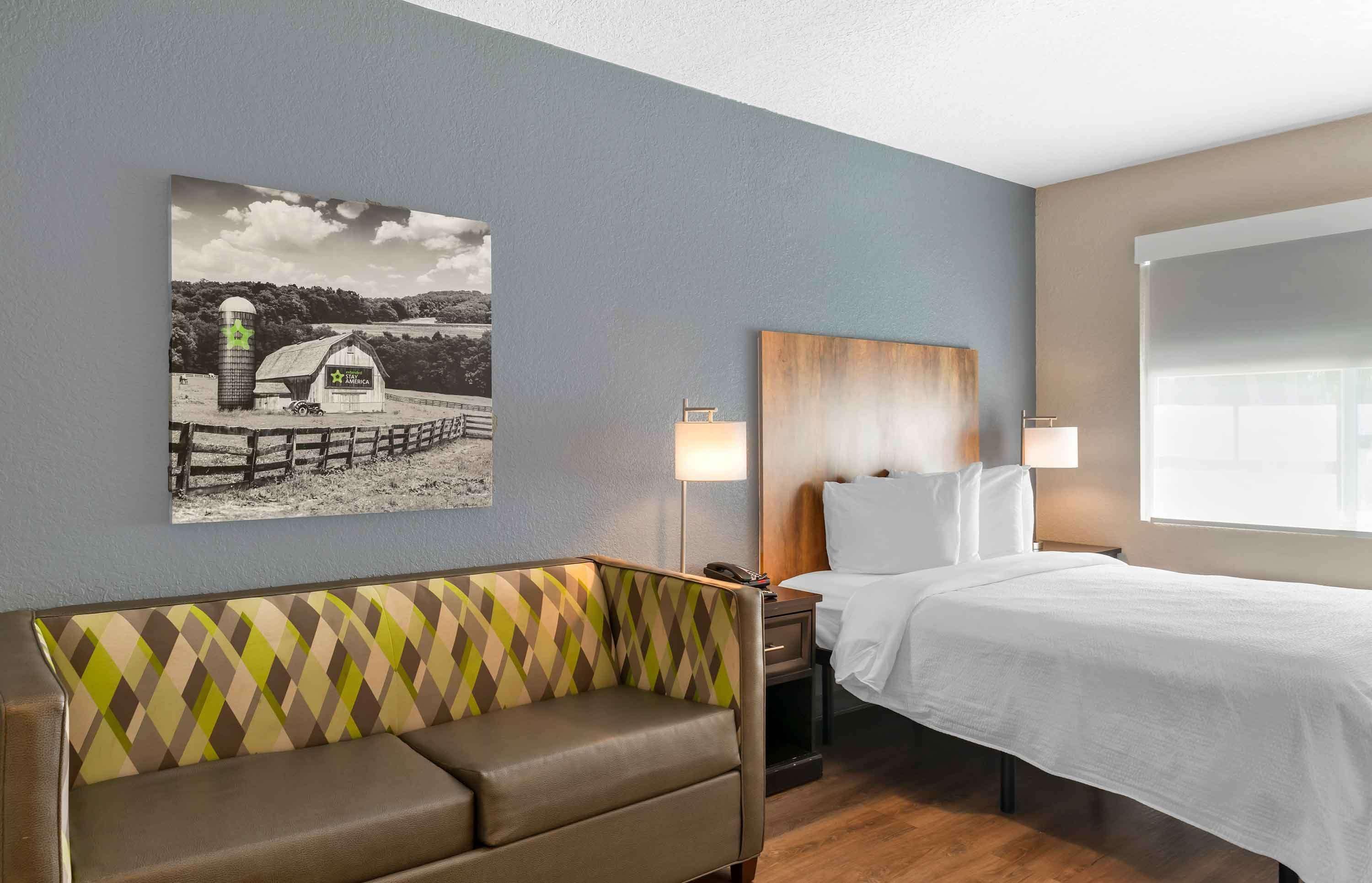 Quarto de hotel com cama, mesa de cabeceira, luminária, sofá e um quadro na parede com a imagem de um celeiro e silo.