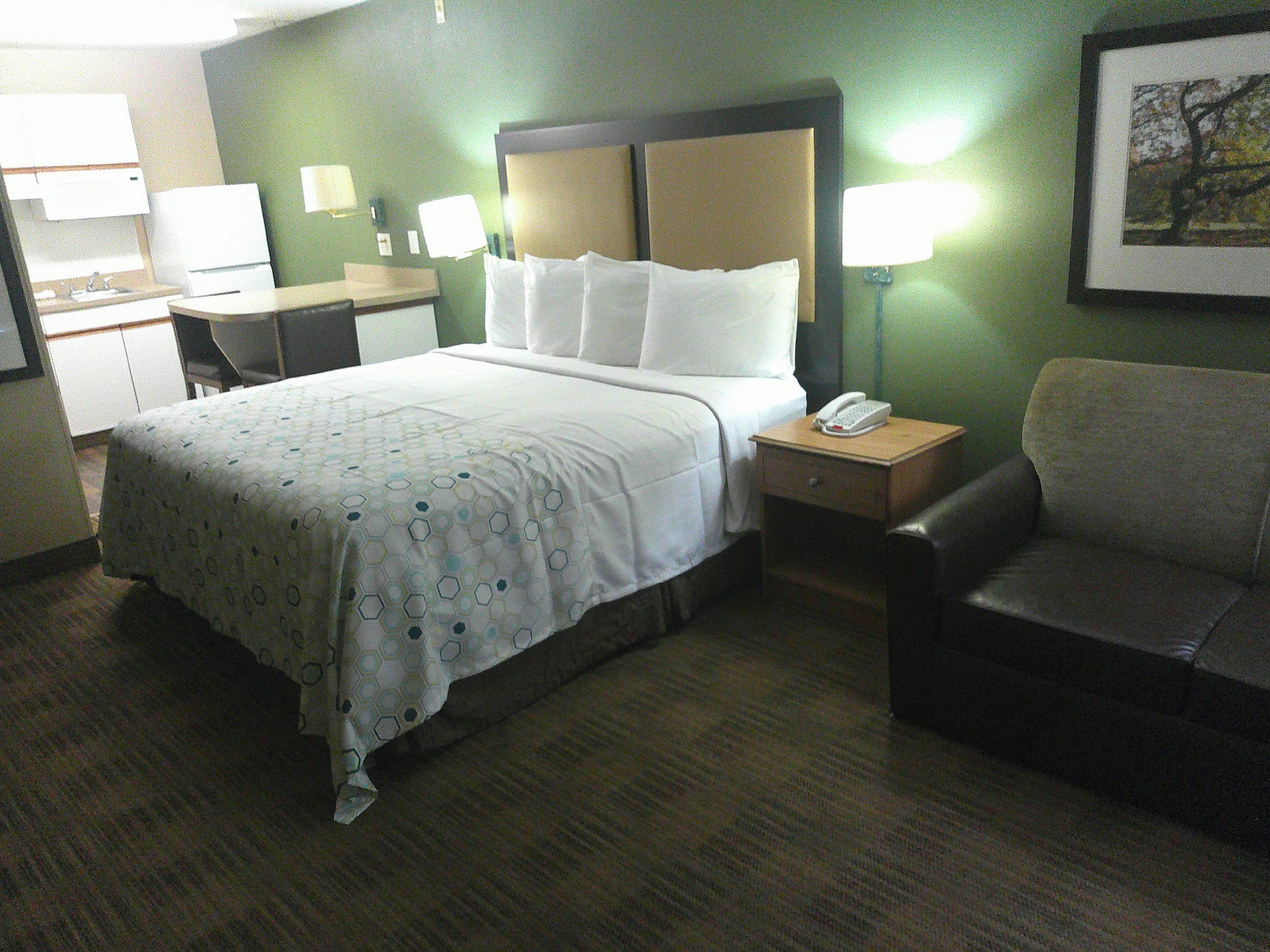 Quarto de hotel com cama, mesa de cabeceira, cadeira e escrivaninha com luminária.
