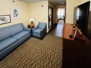 Quarto de hotel com sofá, poltrona, televisão, luminária e obra de arte na parede.