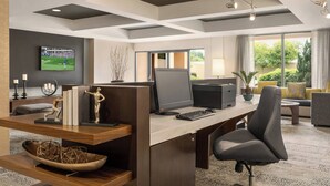 Escritório moderno com mesa, computador e cadeira ergonômica. Há uma televisão na parede e uma janela com vista para o verde.