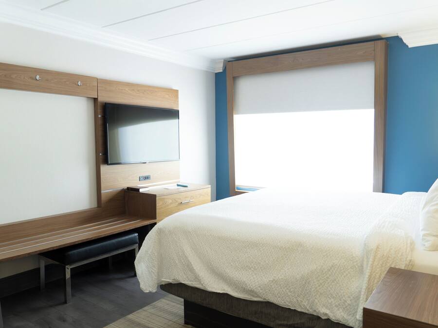 Quarto de hotel com uma cama, escrivaninha, TV de tela plana, janela e cabeceira de madeira.