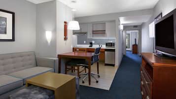 Quarto de hotel com sofá, uma mesa pequena, cozinha compacta, televisão e corredor que dá para outro quarto.