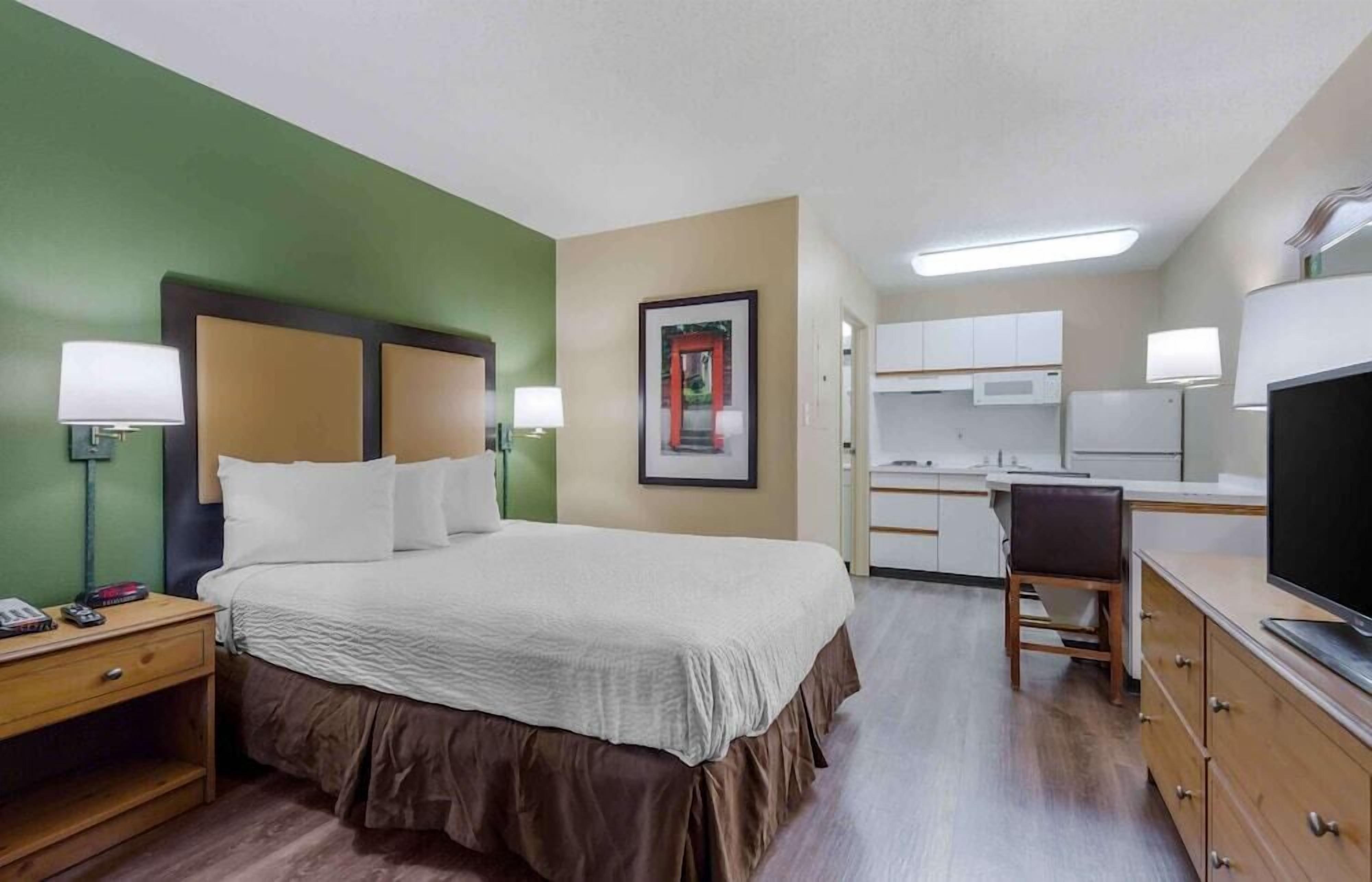 Quarto de hotel com cama, mesa de cabeceira, abajur, escrivaninha, cadeira, cozinha compacta e televisão.