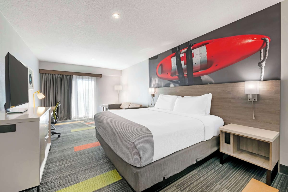Quarto de hotel com uma cama grande, uma mesa, uma cadeira, uma televisão e um quadro de um carro vermelho na parede.