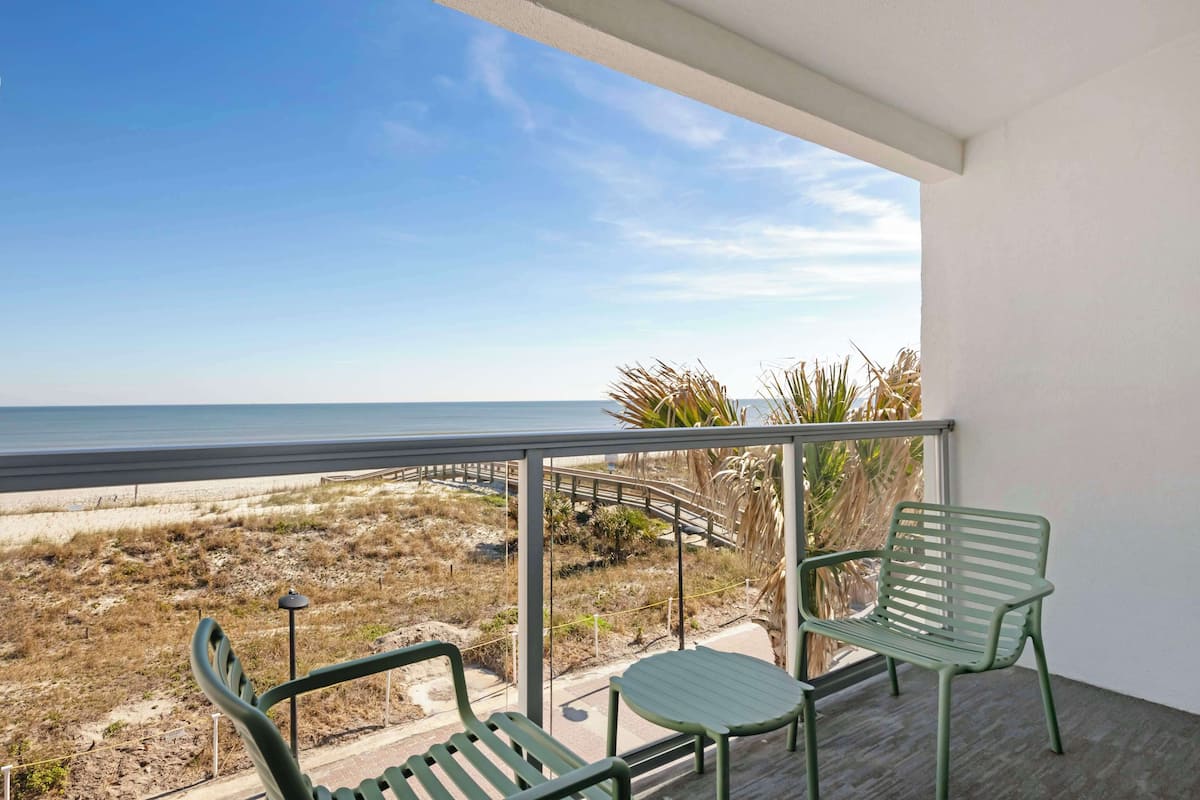 Varanda com duas cadeiras verdes e uma pequena mesa, com vista para uma praia e um calçadão.