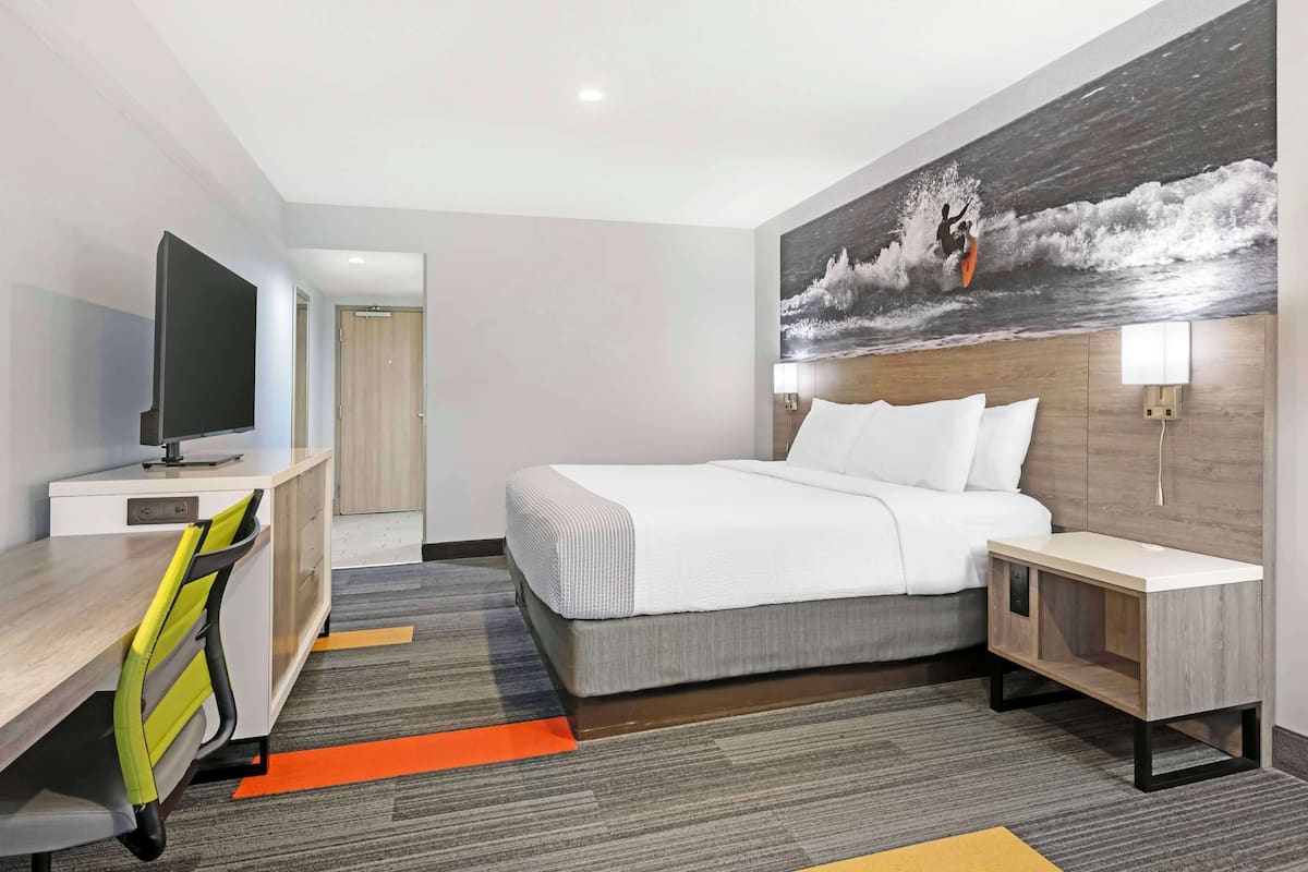 Quarto de hotel moderno com uma cama, uma escrivaninha com televisão de tela plana, uma cadeira e um quadro de um surfista em uma onda.