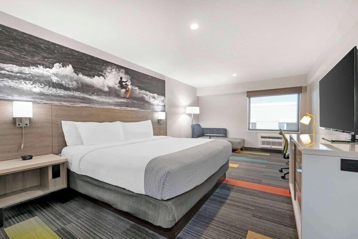 Quarto de hotel com cama, escrivaninha, televisão e um quadro de um surfista na onda.