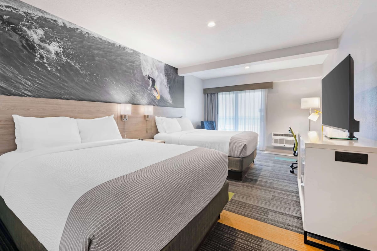 Quarto de hotel com duas camas, um quadro abstrato grande na parede, uma mesa com computador e uma janela com cortinas.