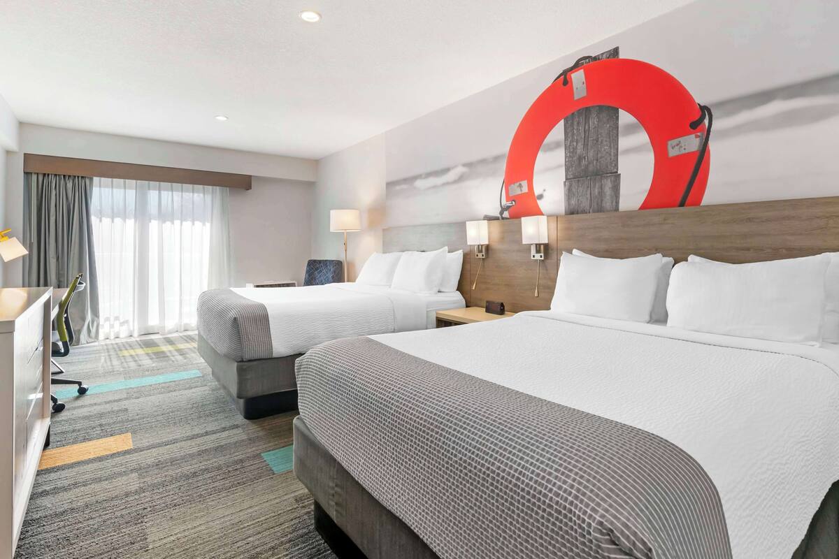 Quarto de hotel com duas camas, uma escrivaninha, uma cadeira, um abajur e uma grande decoração circular na parede.