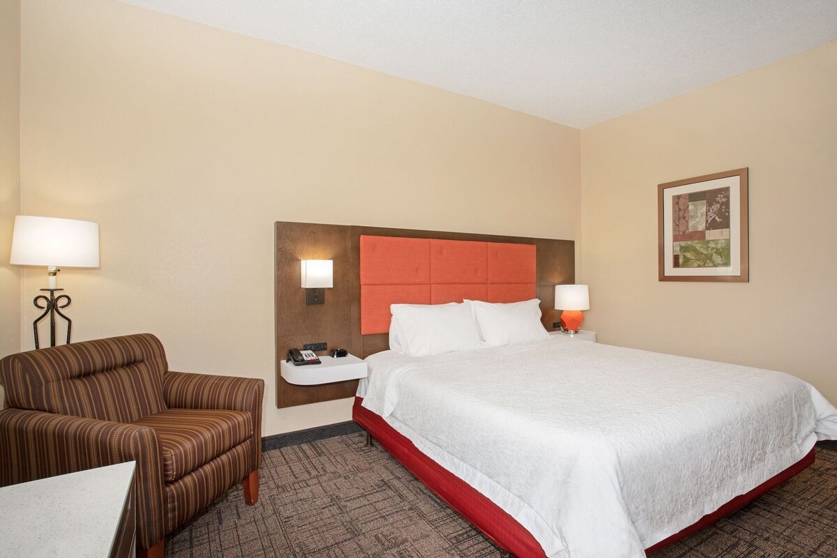 Quarto de hotel com cama, poltrona, escrivaninha, mesa de cabeceira e luminária fixada na parede.