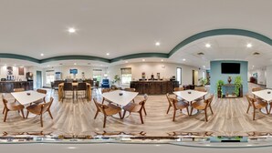 Uma sala de jantar ampla e bem iluminada, com várias mesas e cadeiras, uma cozinha ao fundo e decoração de interiores moderna.