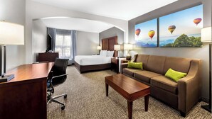Quarto de hotel com cama, sofá, escrivaninha e televisão. Há três quadros na parede e uma janela com cortinas.
