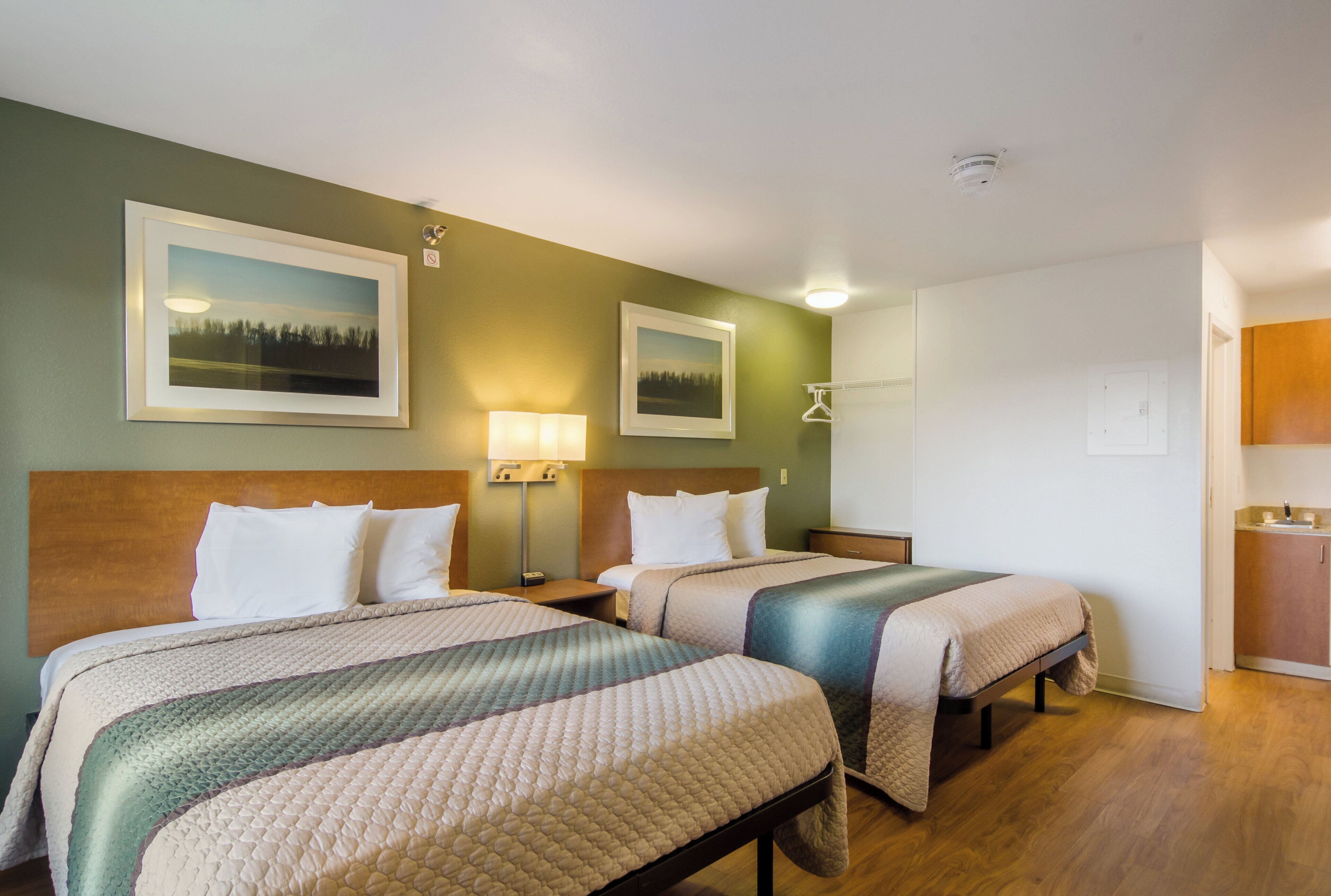Quarto de hotel com duas camas, uma mesa de cabeceira com abajur, uma luminária na parede e uma porta para o banheiro.