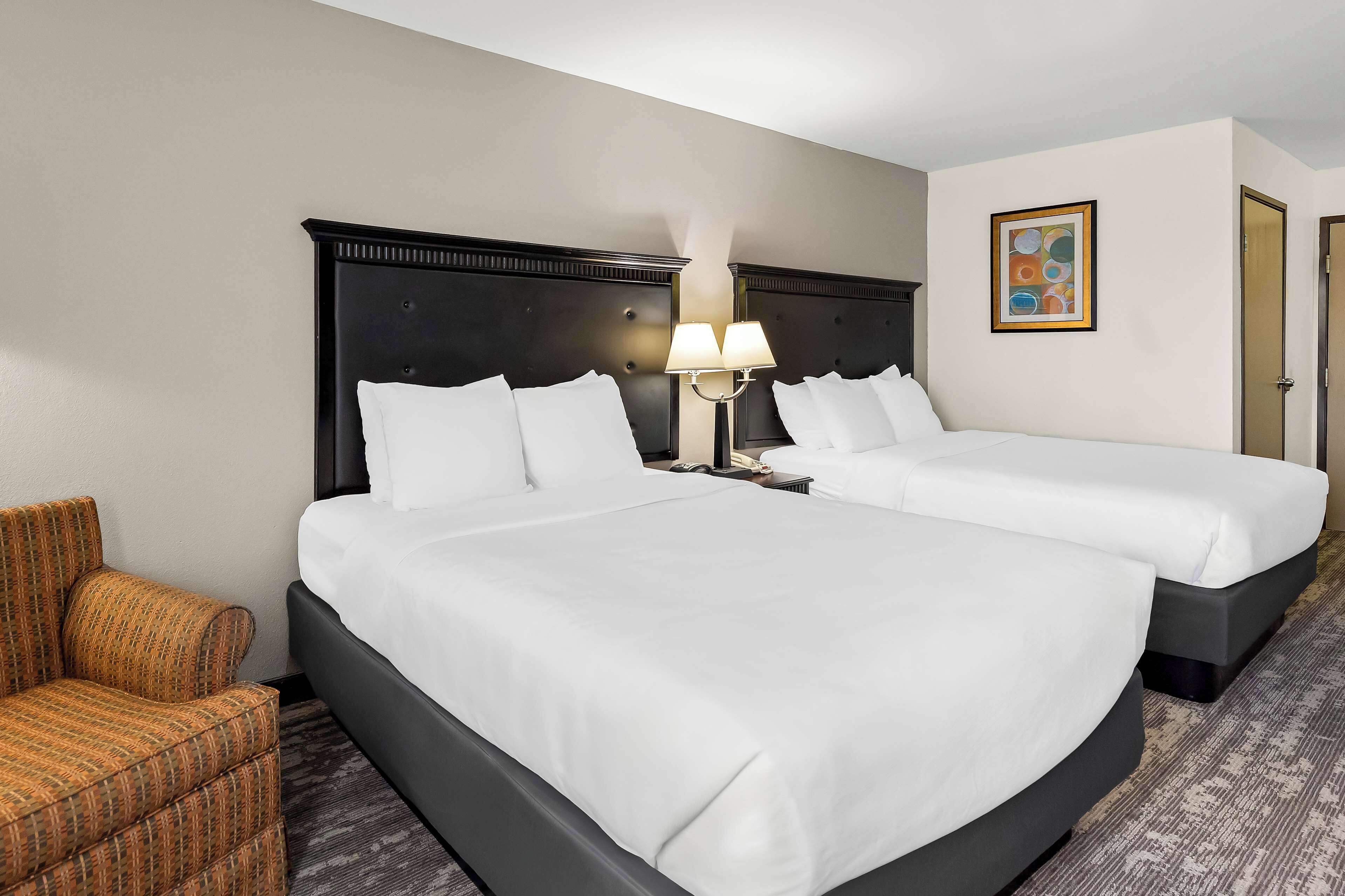 Quarto de hotel com duas camas, uma cadeira, um abajur e um quadro na parede.