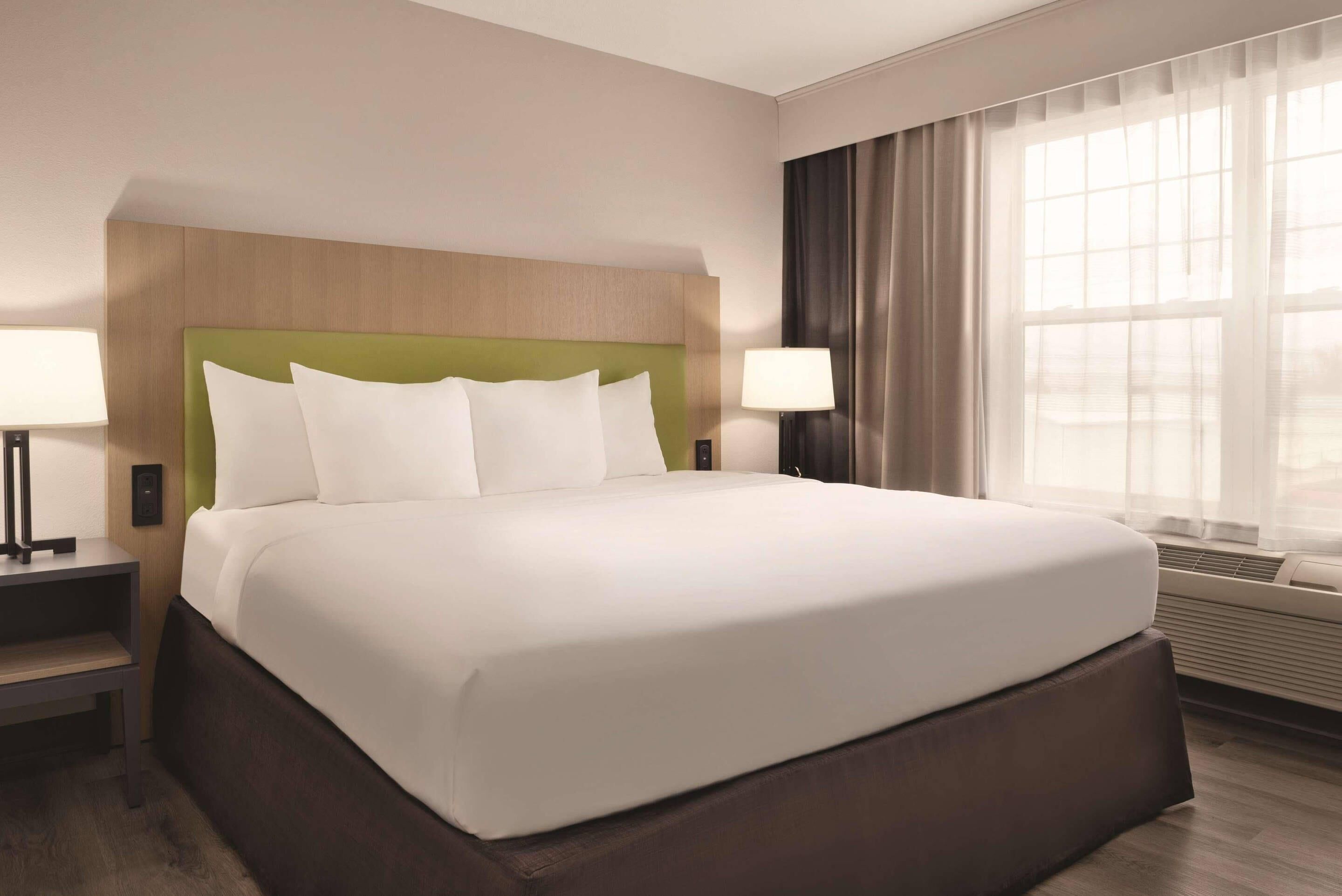 Quarto de hotel com uma cama grande, cabeceira de madeira, duas mesas de cabeceira com luminárias e uma janela com cortinas.