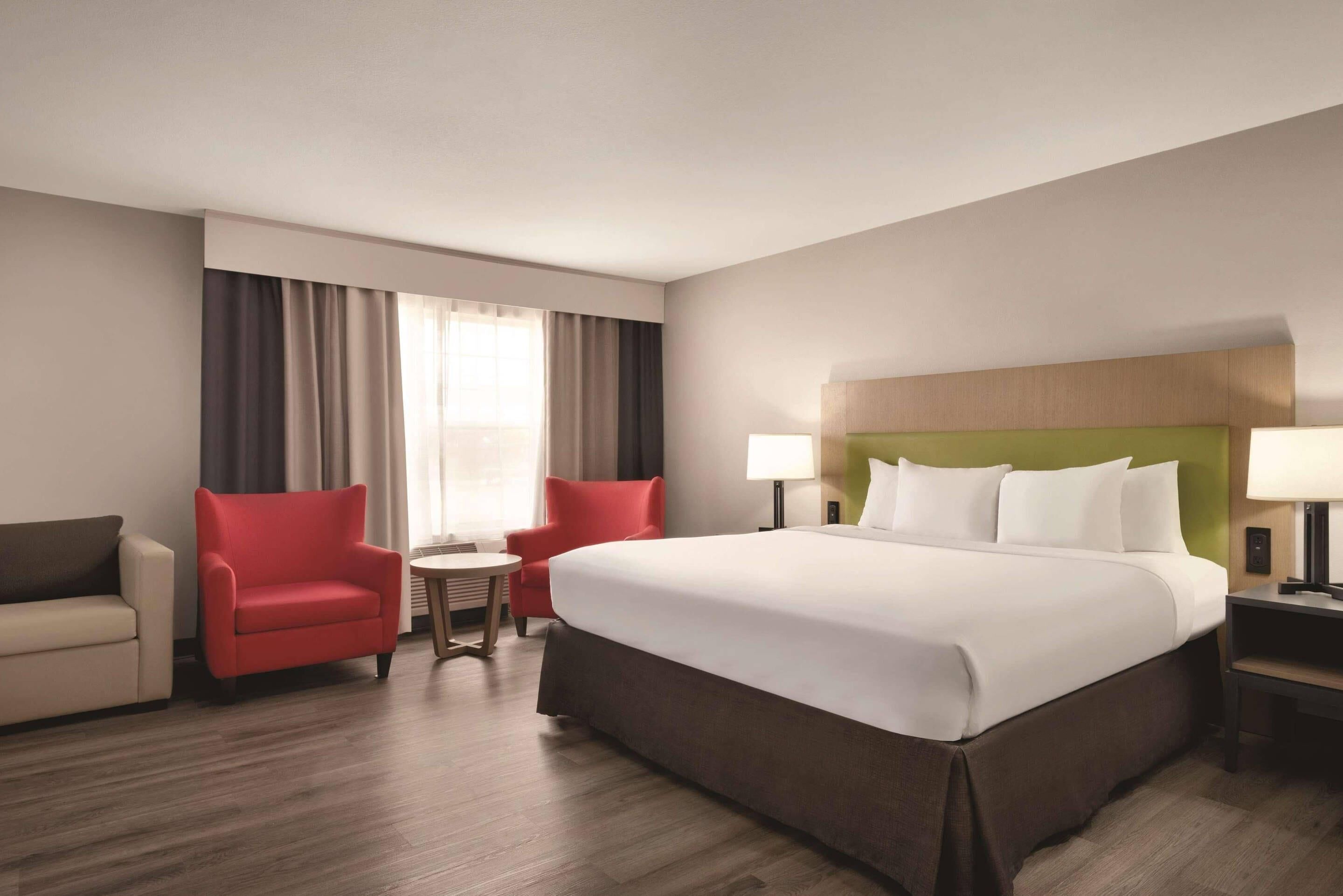Quarto de hotel com uma cama grande, duas poltronas vermelhas, uma mesinha e uma luminária.