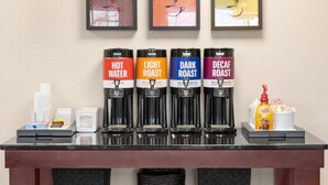 Um espaço para café com quatro dosadores identificados como 'Água Quente', 'Torra Clara', 'Torra Escura' e 'Descafeinado'. Há pilhas de copos brancos e uma máquina de café sobre a mesa.
