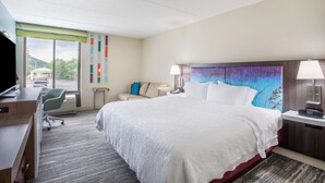 Quarto de hotel com uma cama grande, uma escrivaninha com computador, uma cadeira, uma mesa de cabeceira com abajur e uma janela com vista para o exterior.