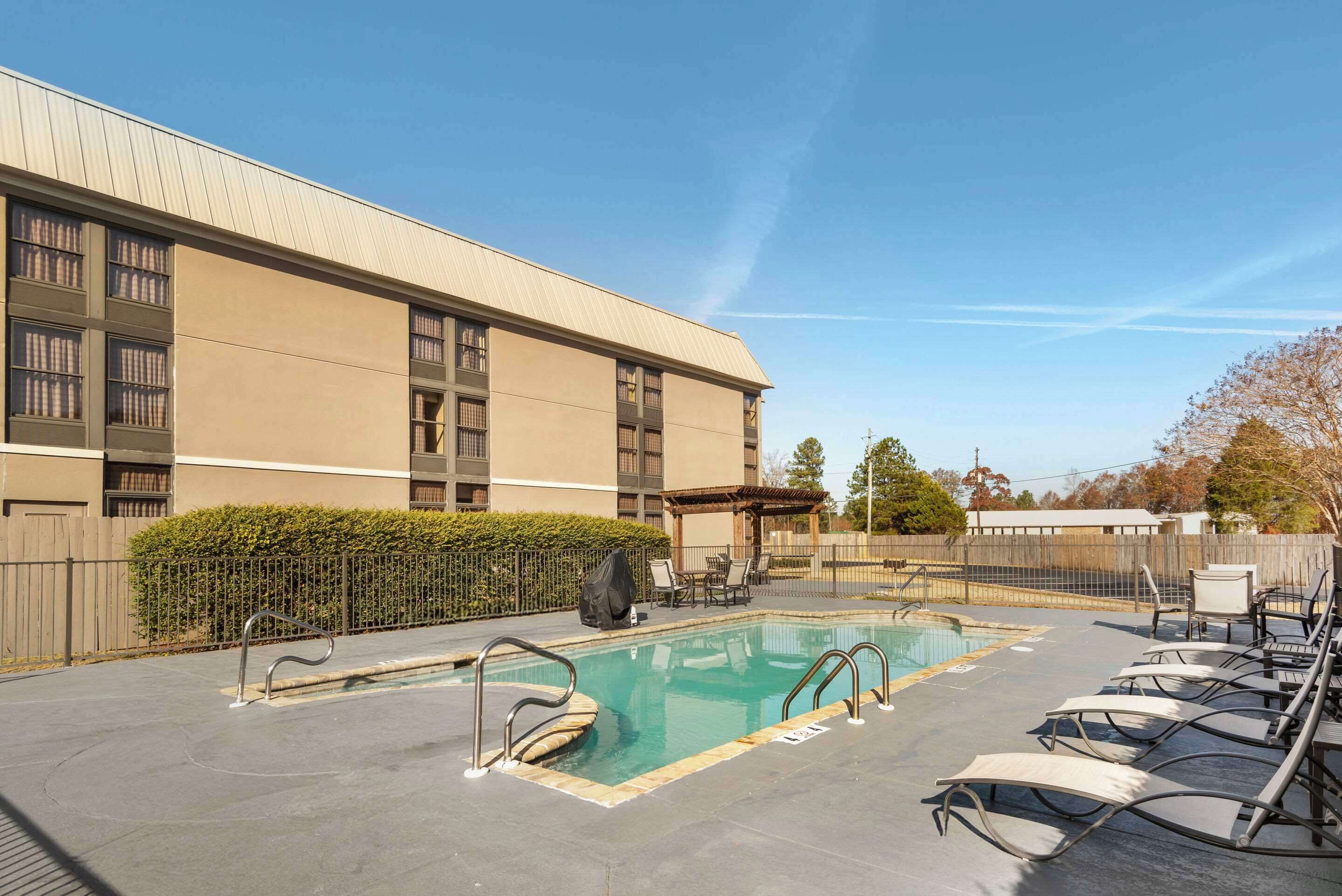 Piscina com espreguiçadeiras, área de estar coberta e edifício com várias janelas e telhado plano.