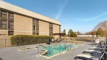 Piscina com espreguiçadeiras, área de estar coberta e edifício com várias janelas e telhado plano.