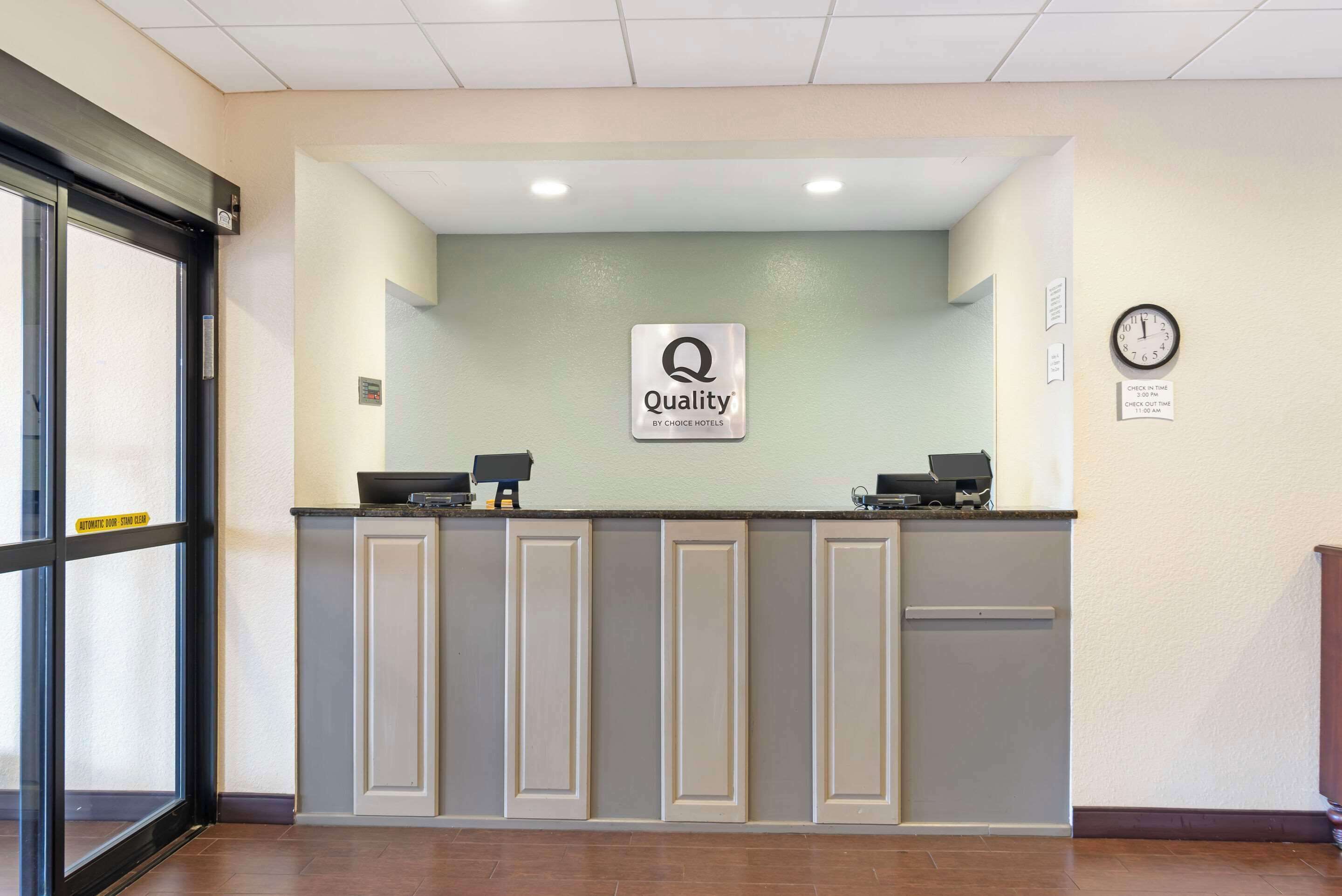 Uma mesa de recepção com uma placa indicando "Quality by Choice Hotels", um relógio e uma placa com a inscrição "Horário de Check-in".
