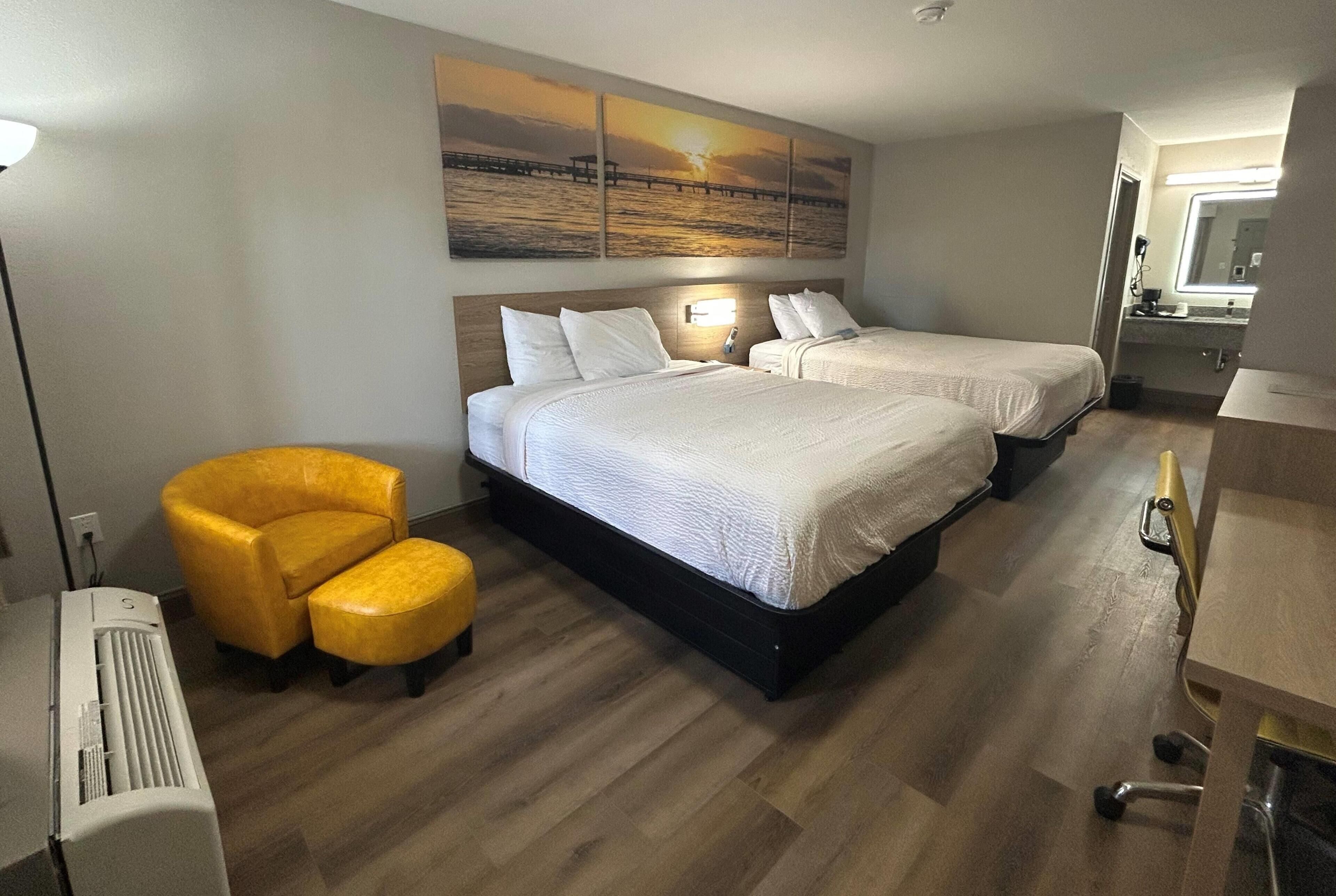 Quarto de hotel com duas camas, uma cadeira, uma escrivaninha e um quadro grande na parede.