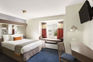 Quarto de hotel com cama, escrivaninha, cadeira, televisão fixada na parede e janela com cortinas.