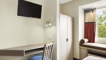 Quarto de hotel com cama, escrivaninha com cadeira, televisão na parede, guarda-roupa e janela com cortinas.