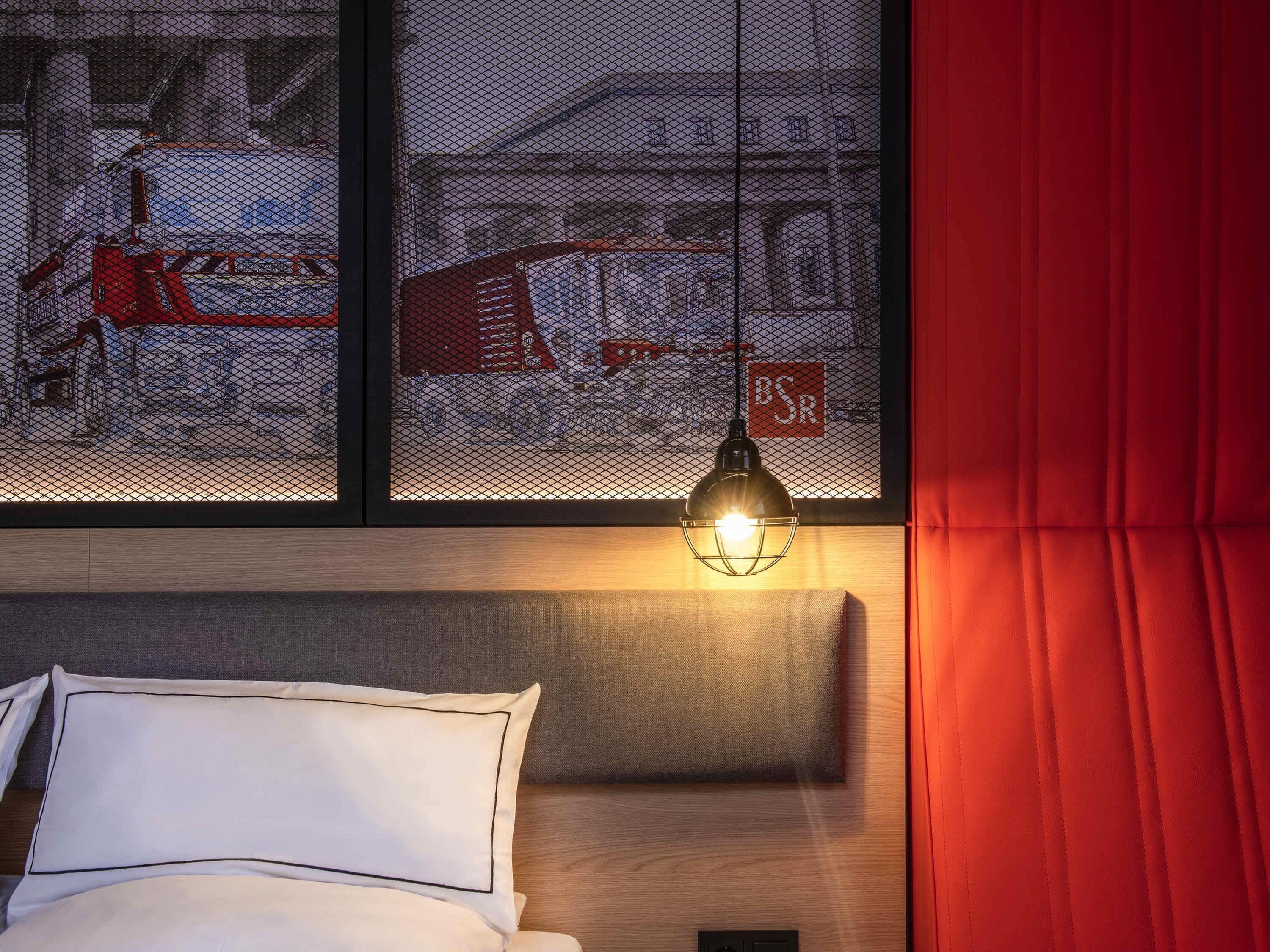 Cama com roupa branca, cabeceira e janela com vista para um caminhão vermelho e prédios.